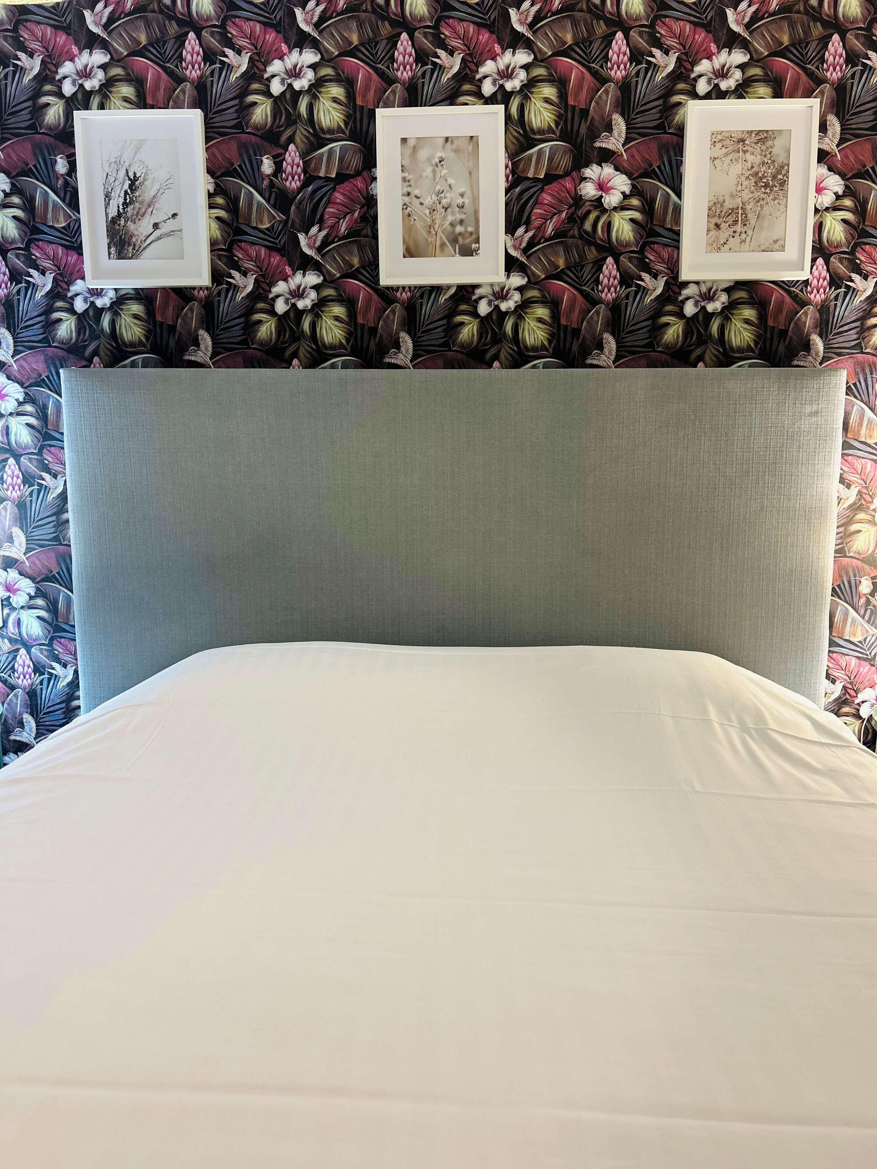 Cama com cabeceira cinza e roupa de cama branca, com papel de parede floral e três quadros emoldurados.