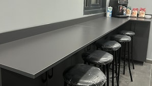 Uma área de bar com um balcão comprido, banquetas pretas, uma máquina de café e lanches embalados.