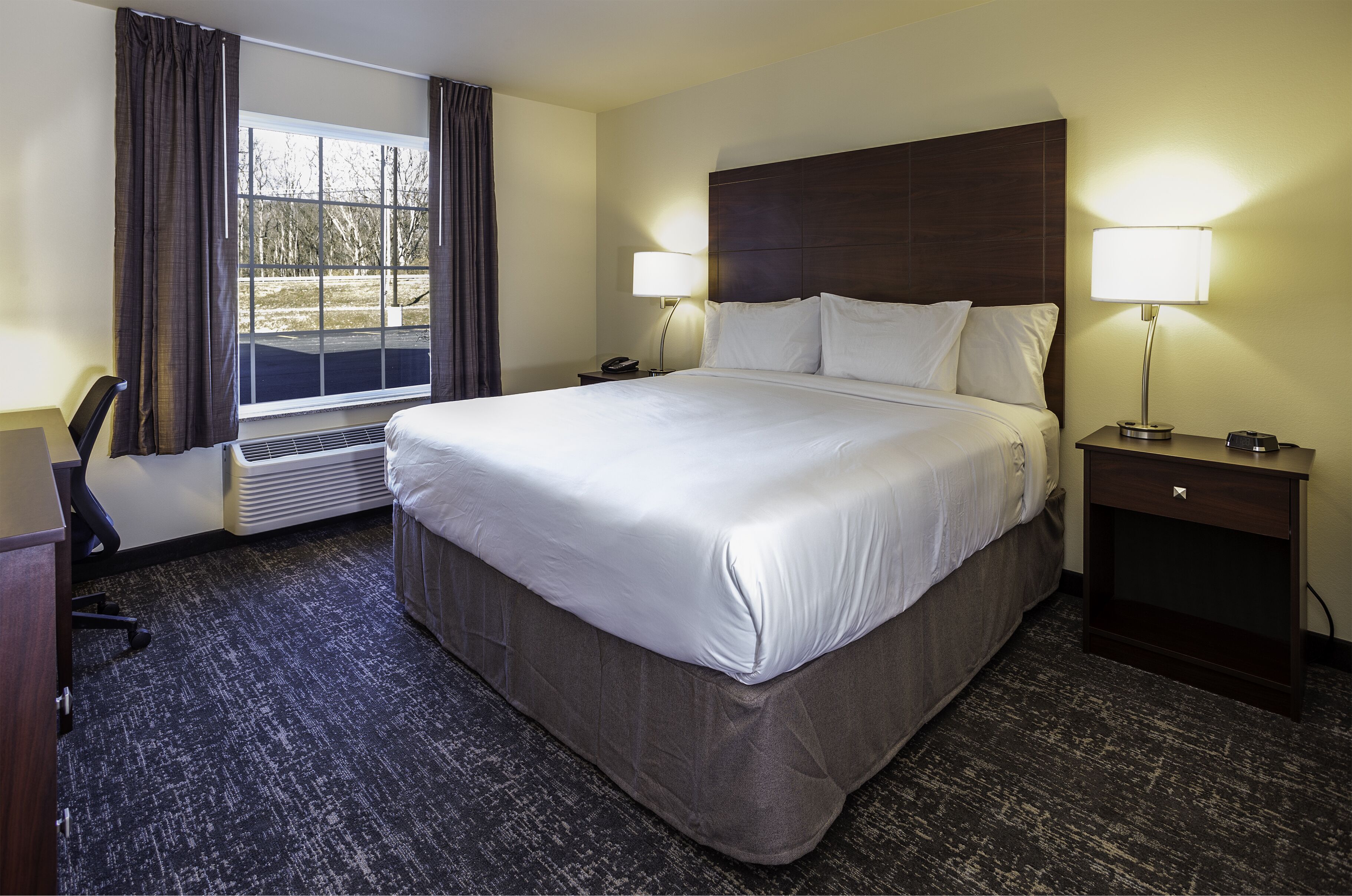 Quarto de hotel com uma cama grande, duas luminárias de cabeceira, uma mesa de trabalho com telefone e uma janela com cortinas.