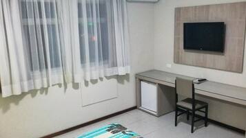 Quarto pequeno e limpo, com escrivaninha, cadeira e TV fixada na parede.