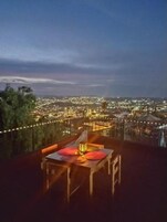 Mesa com luminária e almofadas em varanda com vista para a cidade à noite.