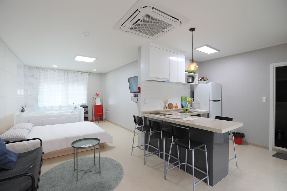 Quarto de hotel moderno com cozinha compacta, cama com edredom branco, mesa redonda pequena e tapete cinza.