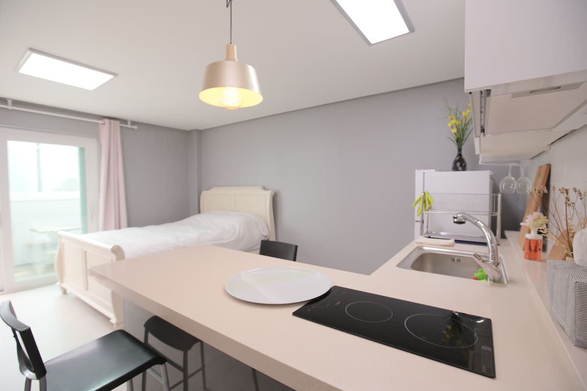 Quarto de hotel moderno com cozinha compacta, cama, escrivaninha e uma janela grande.