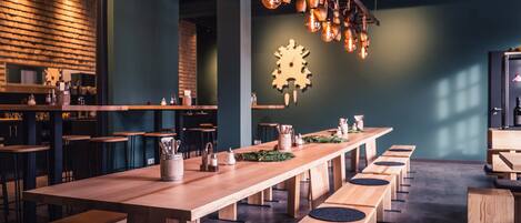 Restaurante moderno com longas mesas e bancos de madeira, panelas de cobre penduradas e uma parede verde com decoração de engrenagem de madeira.