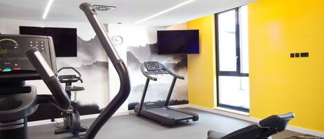 Uma academia com equipamentos de exercícios, esteira e televisão presa na parede.