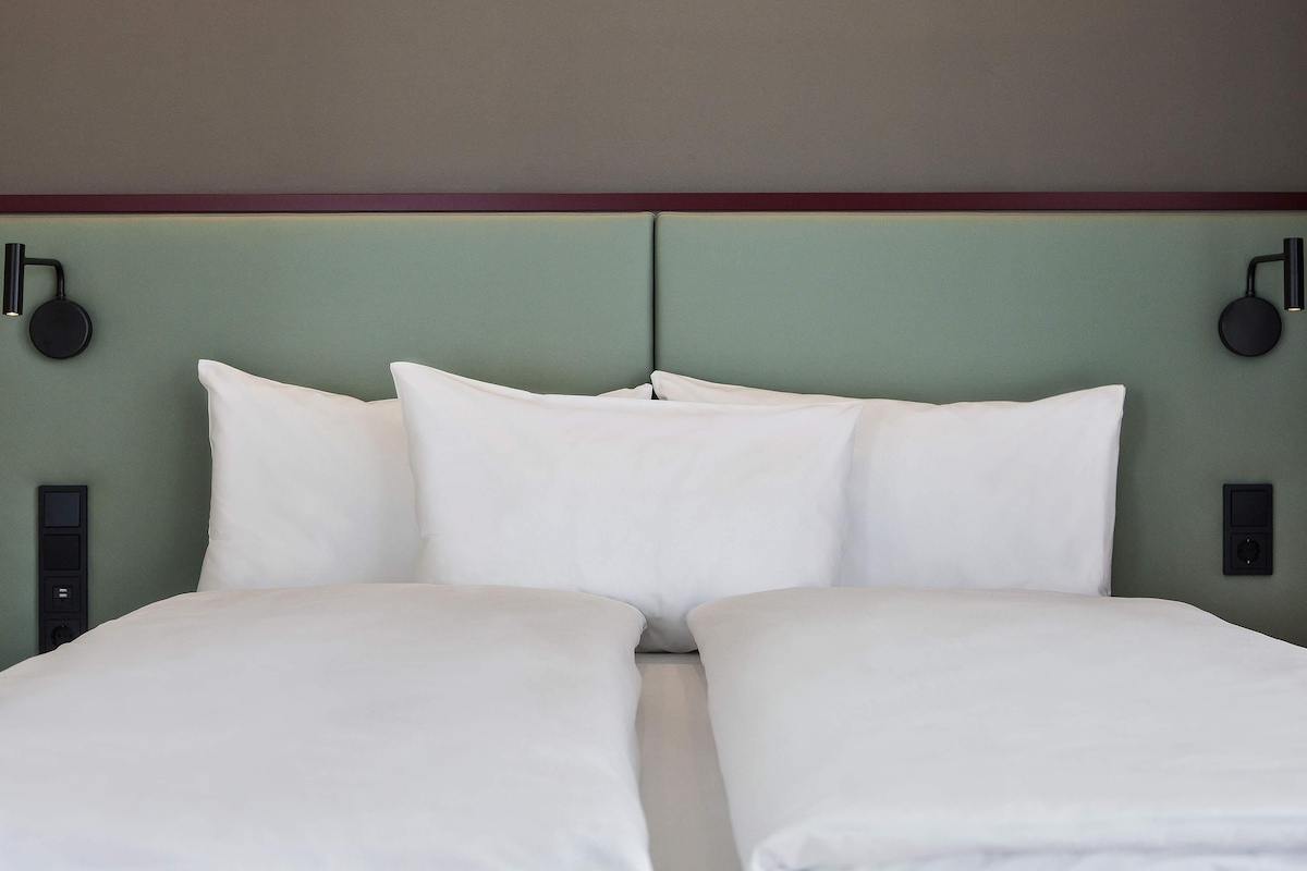 Cama de casal com roupa de cama branca, cabeceira verde e duas luminárias fixadas na parede.