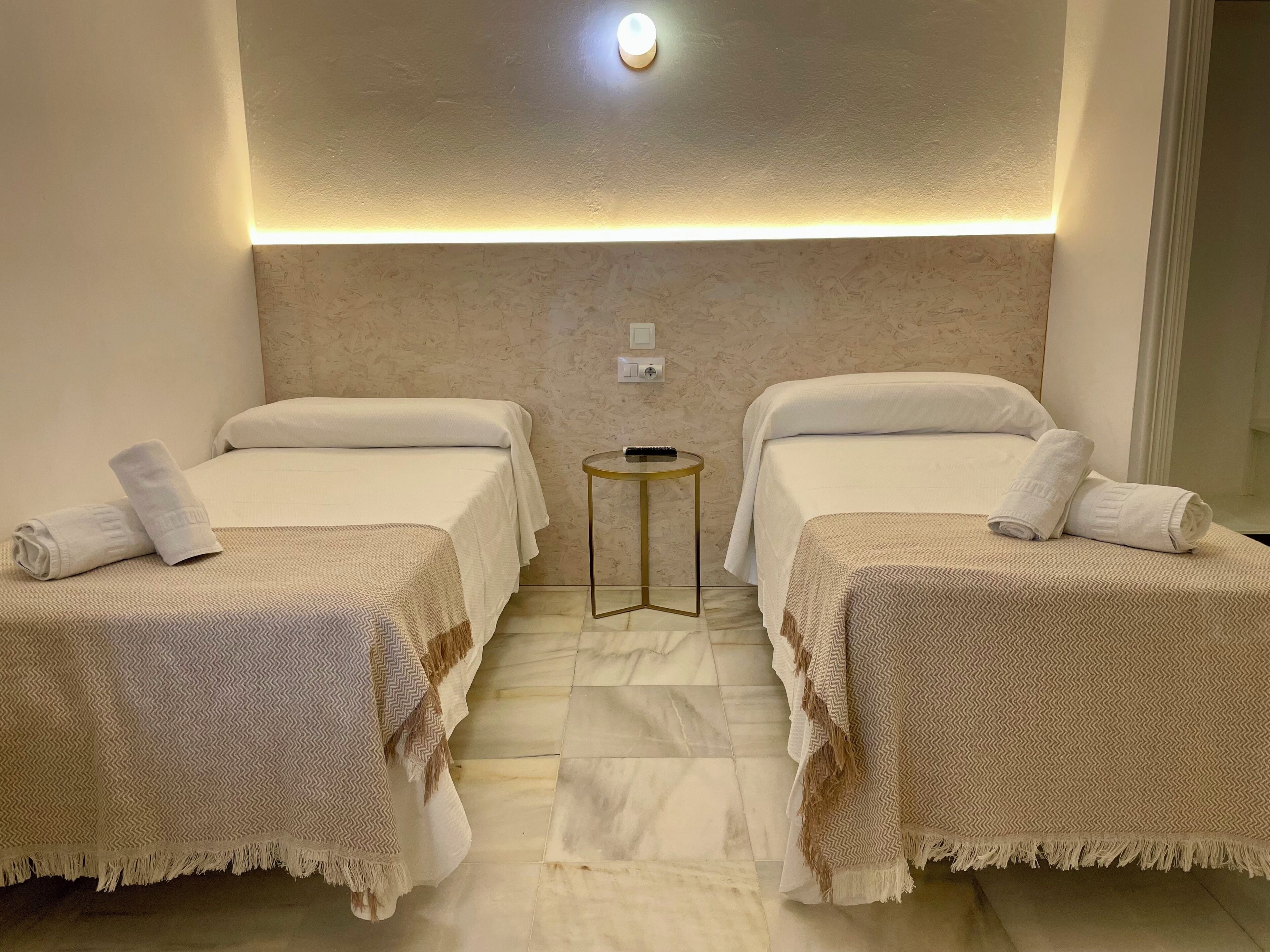 Quarto de hotel com duas camas, uma mesa redonda pequena e uma luminária fixada na parede.