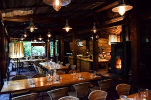 Interior rústico de restaurante com mesas de madeira, cadeiras de vime, luminárias pendentes, parede de tijolos e lareira.