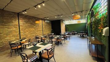 Restaurante com paredes de tijolo, mesas e cadeiras de madeira. Há luminárias pendentes e uma parede verde com plantas.