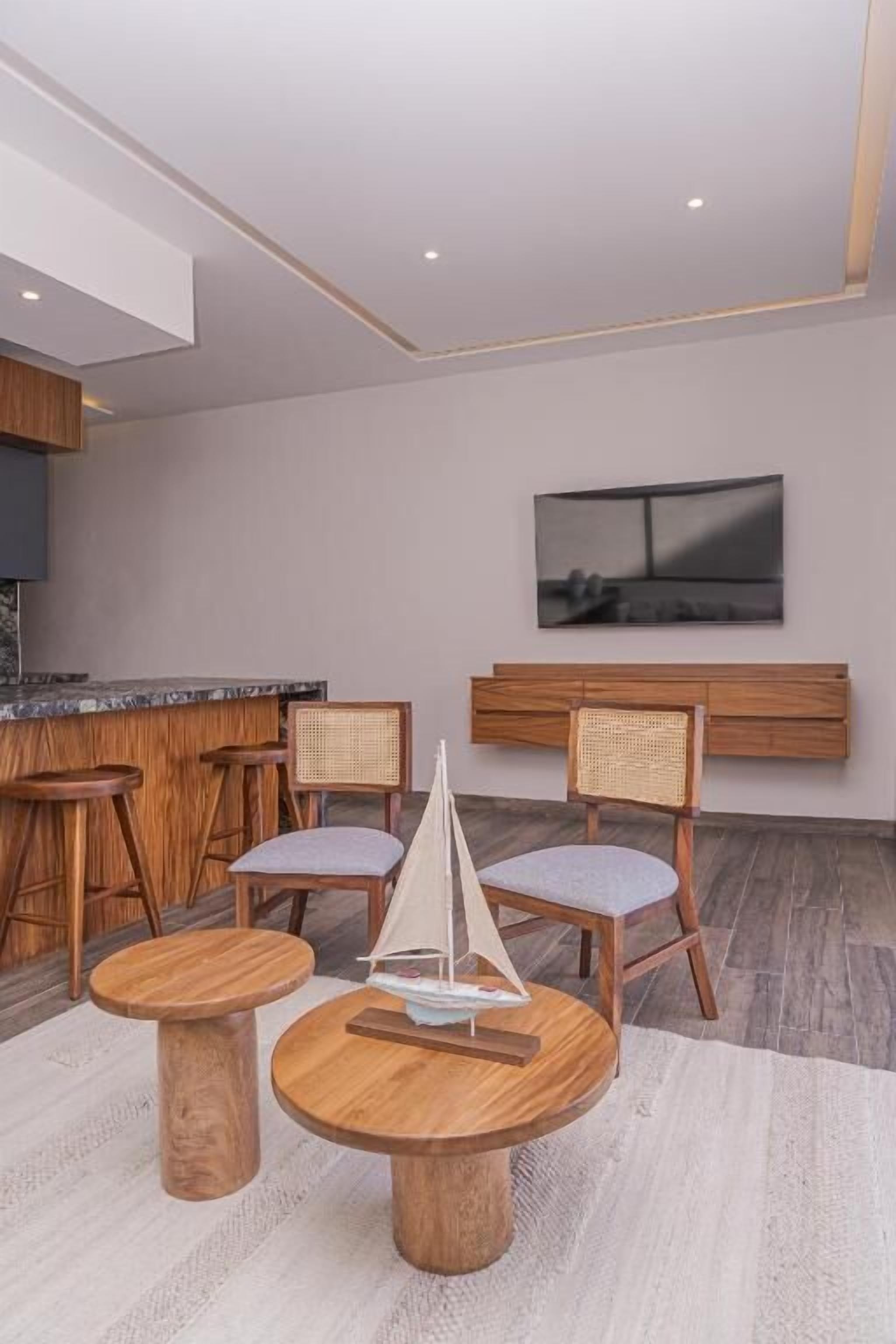 Um interior moderno com uma mesa de madeira redonda, dois banquetas de bar, uma miniatura de veleiro e uma televisão fixada na parede.