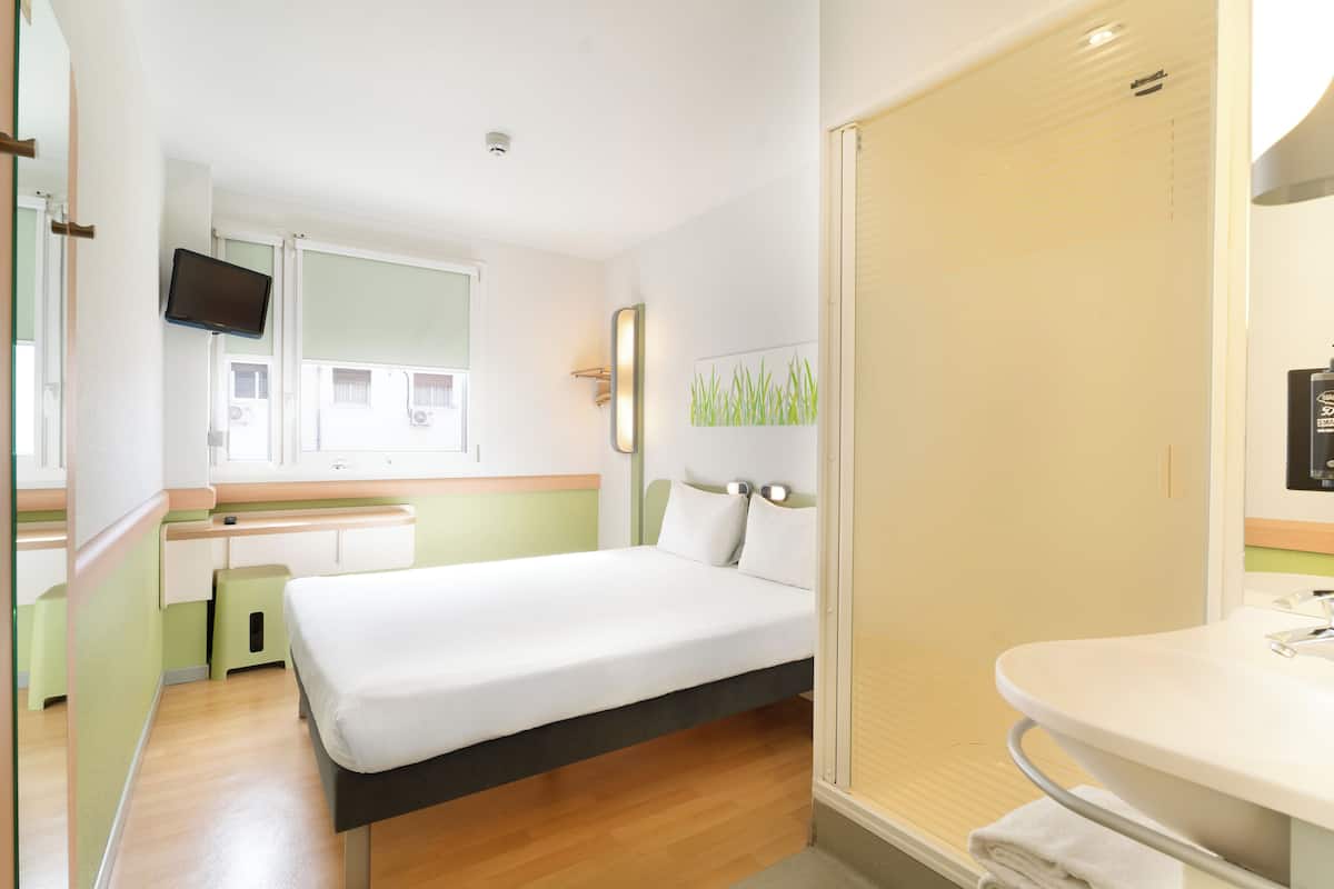 Quarto de hotel com cama, escrivaninha, televisão, banheiro com pia e janela.