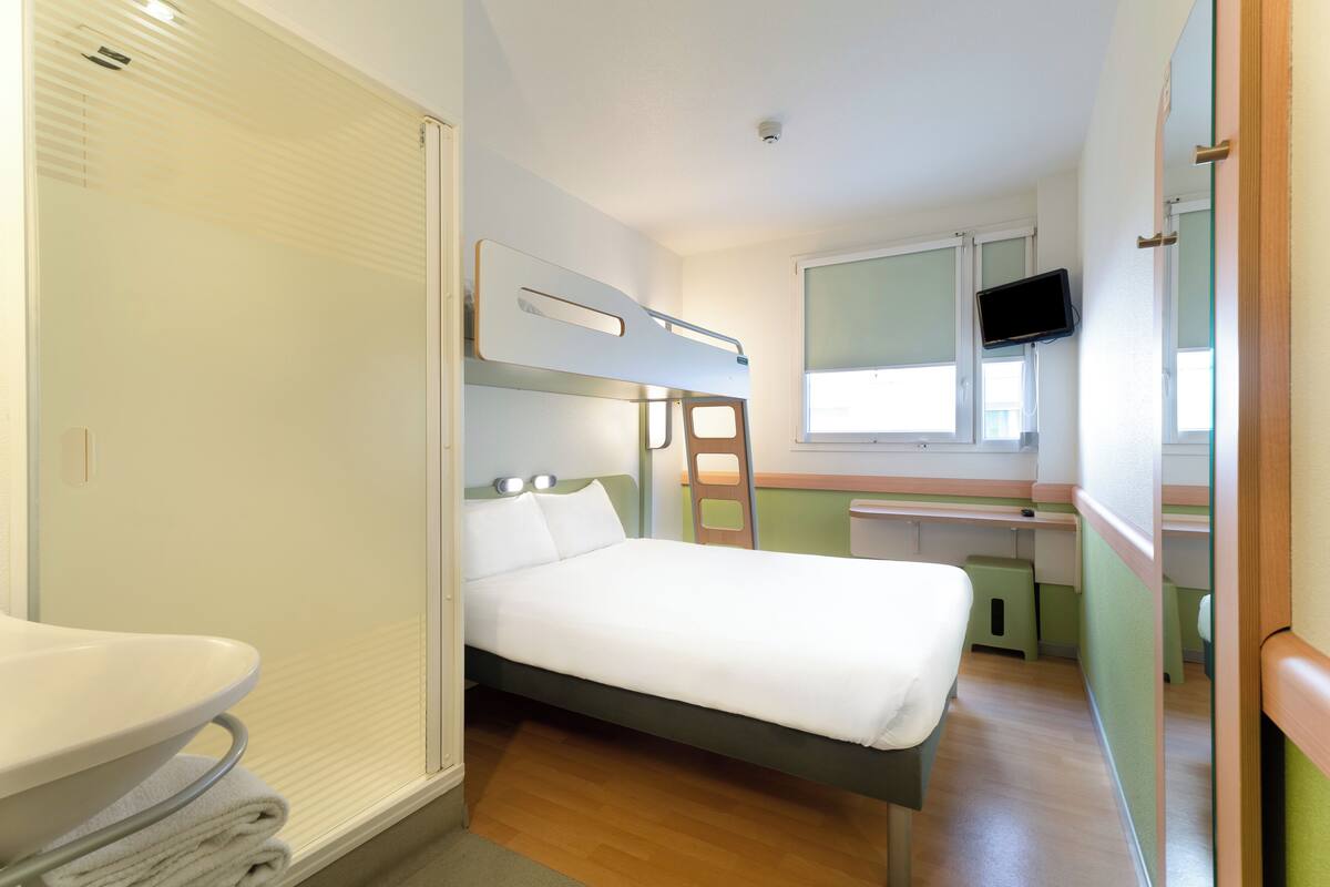 Quarto de hotel compacto com uma cama beliche, uma escrivaninha pequena, um espelho e uma televisão.