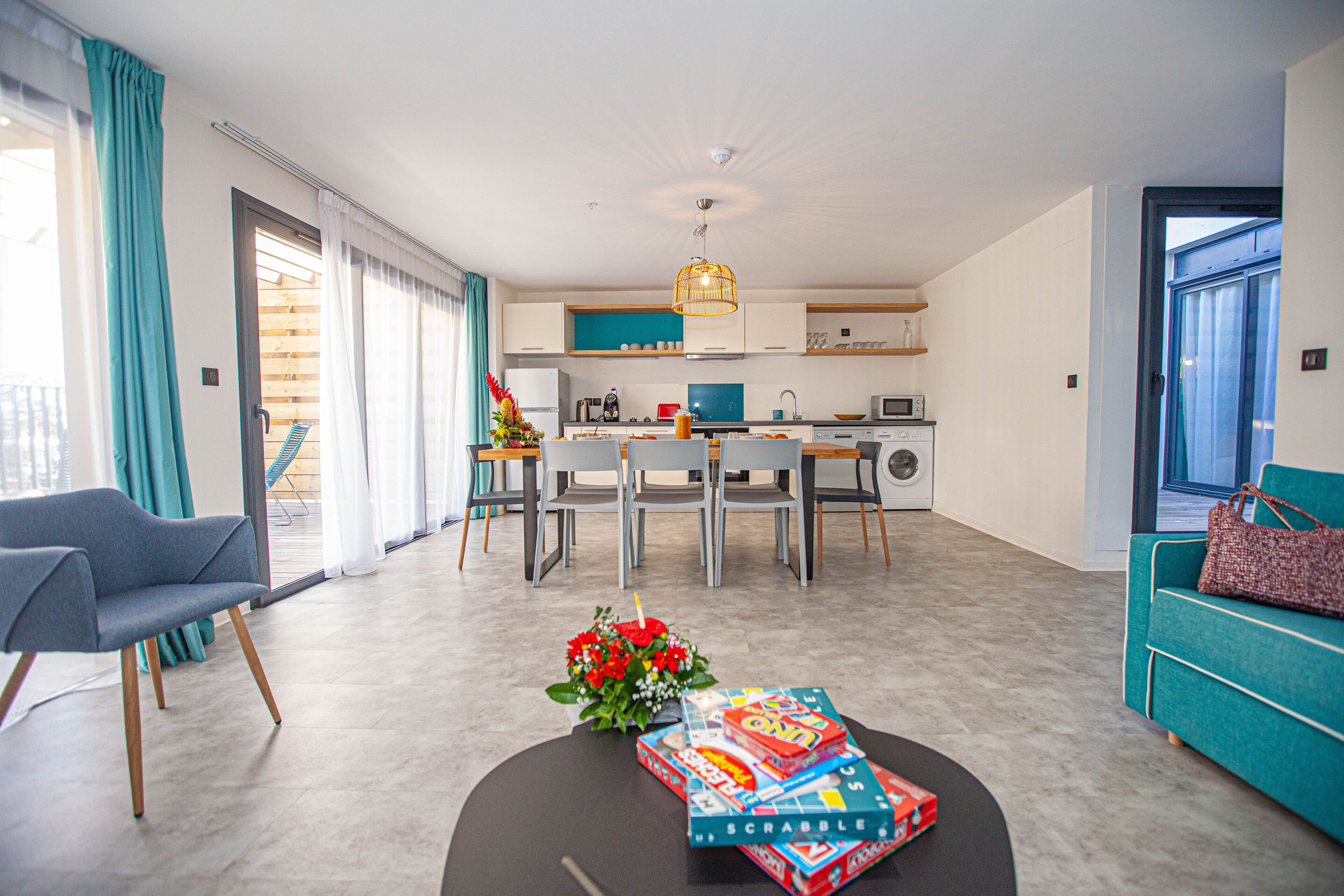 Uma sala de estar moderna com um sofá, uma mesa de centro com um jogo de Scrabble, uma área de jantar com cadeiras e uma cozinha ao fundo.