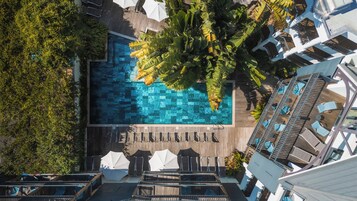 Vista aérea de uma piscina no terraço com cadeiras de sol e guarda-sóis ao redor, cercada por vegetação densa e edifícios vizinhos.