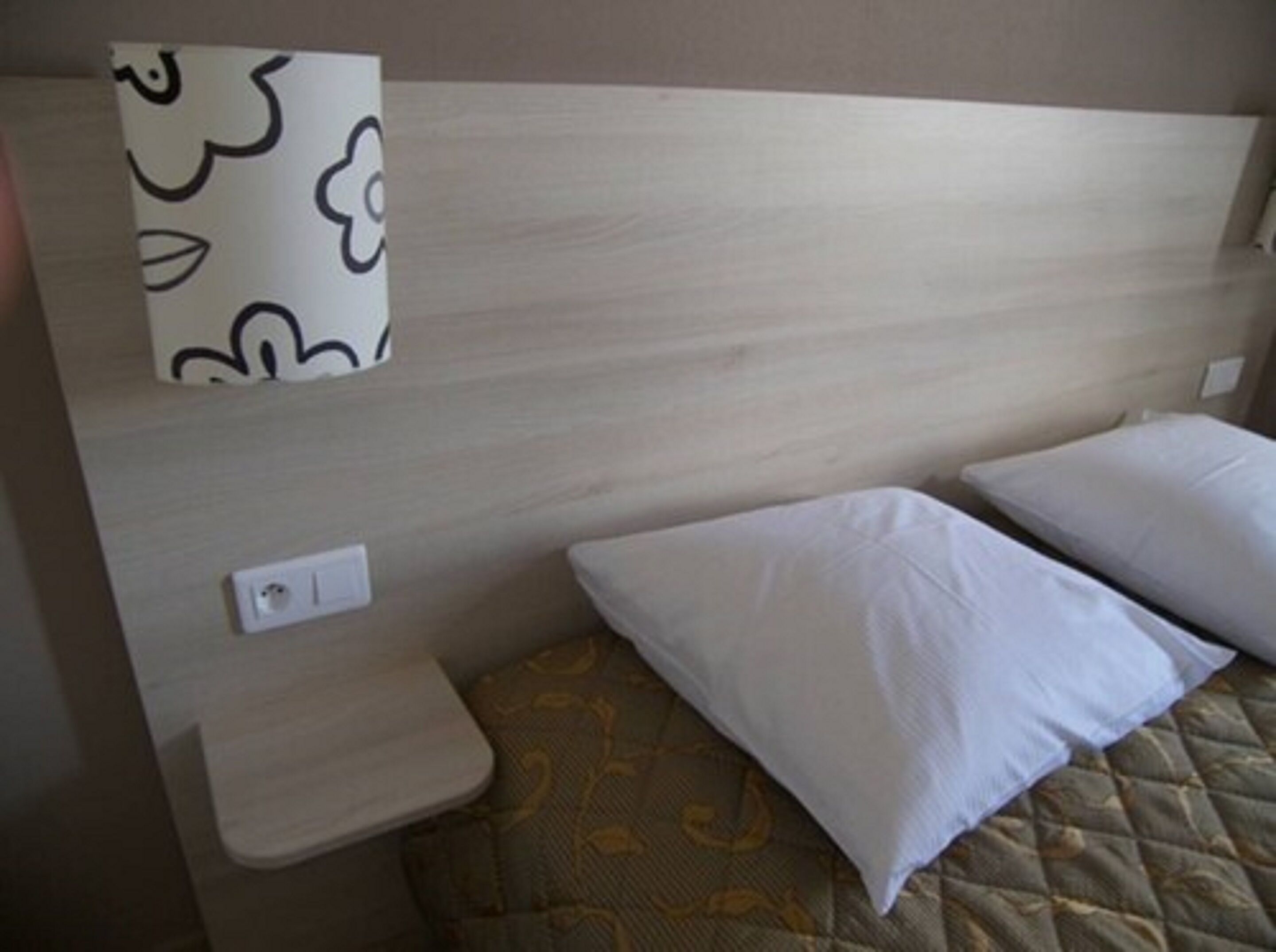 Quarto de hotel com cabeceira de madeira, mesa de cabeceira, duas almofadas e colcha estampada.