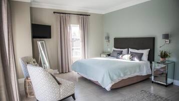 Quarto com cama grande, uma cadeira, televisão fixada na parede e mesa de cabeceira com abajur.