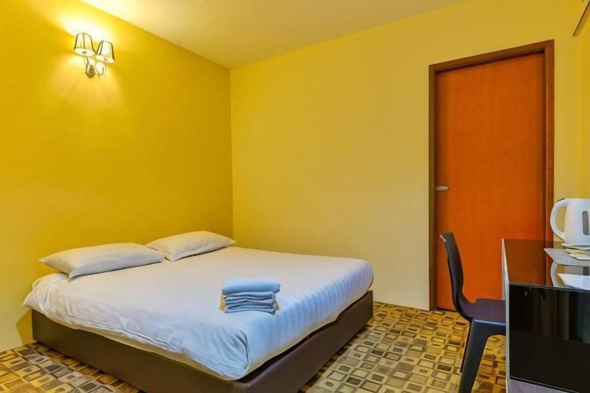 Uma cama de solteiro com colchão e travesseiros brancos, uma mesa pequena com uma cadeira, uma porta e um luminário fixado na parede.