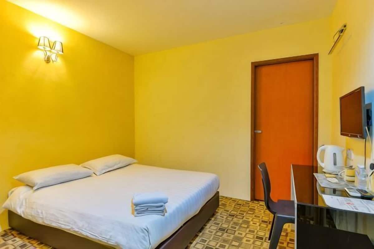 Quarto de hotel com uma cama, escrivaninha, cadeira, televisão e porta.