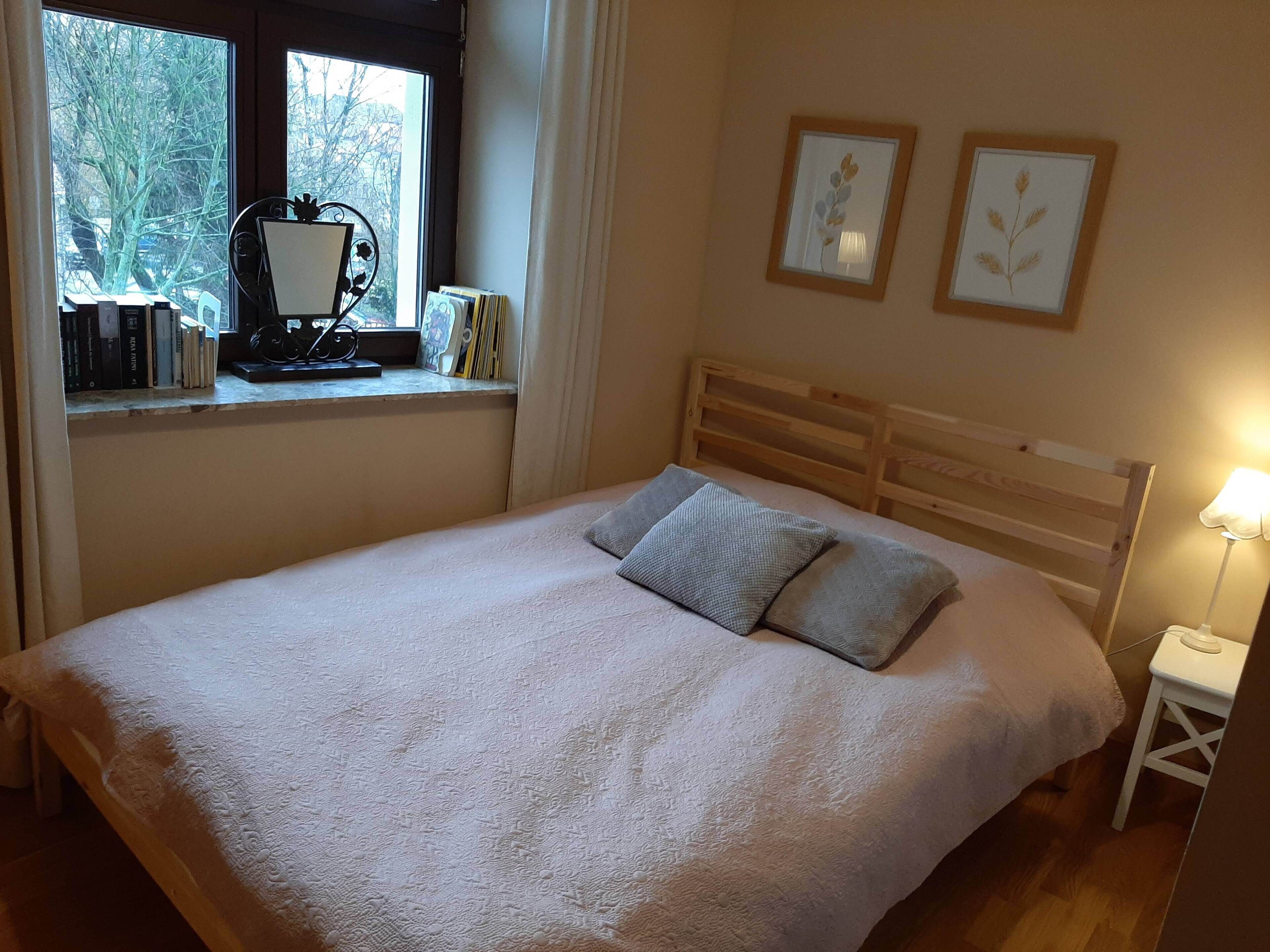 Cama bem arrumada com edredom branco e duas almofadas, cabeceira de madeira, mesa de cabeceira com abajur e janela com vista para árvores e um carro estacionado.