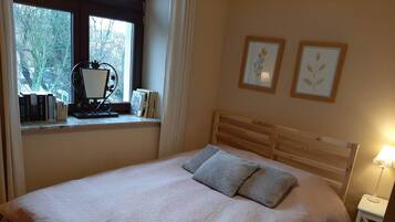 Cama bem arrumada com edredom branco e duas almofadas, cabeceira de madeira, mesa de cabeceira com abajur e janela com vista para árvores e um carro estacionado.