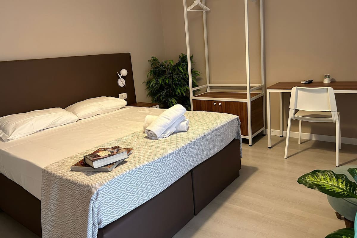 Uma cama bem arrumada com edredom branco e almofadas, uma mesa de cabeceira com luminária, uma pequena escrivaninha com cadeira e uma planta pendente.