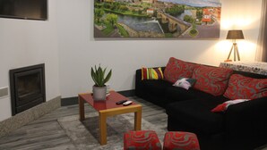 Uma sala de estar com lareira, um sofá com almofadas estampadas, uma mesa de centro com uma planta e um quadro de uma ponte sobre um rio.