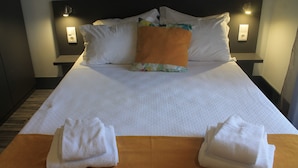 Cama bem arrumada com lençóis brancos e um único travesseiro laranja, dois toalhas dobradas sobre a cama, e luminárias fixadas na parede em ambos os lados.