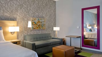 Quarto de hotel com cama, sofá, mesa pequena, cadeira, espelho e um quadro na parede.