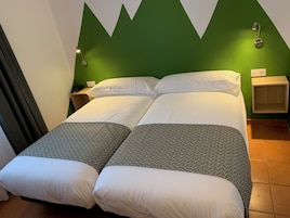 Cama de casal com roupa de cama branca e almofadas estampadas, mesa de cabeceira com abajur e parede verde decorativa.