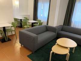 Quarto de hotel moderno com sofá cinzento, duas mesinhas redondas, cadeiras verdes e uma janela grande com cortinas.
