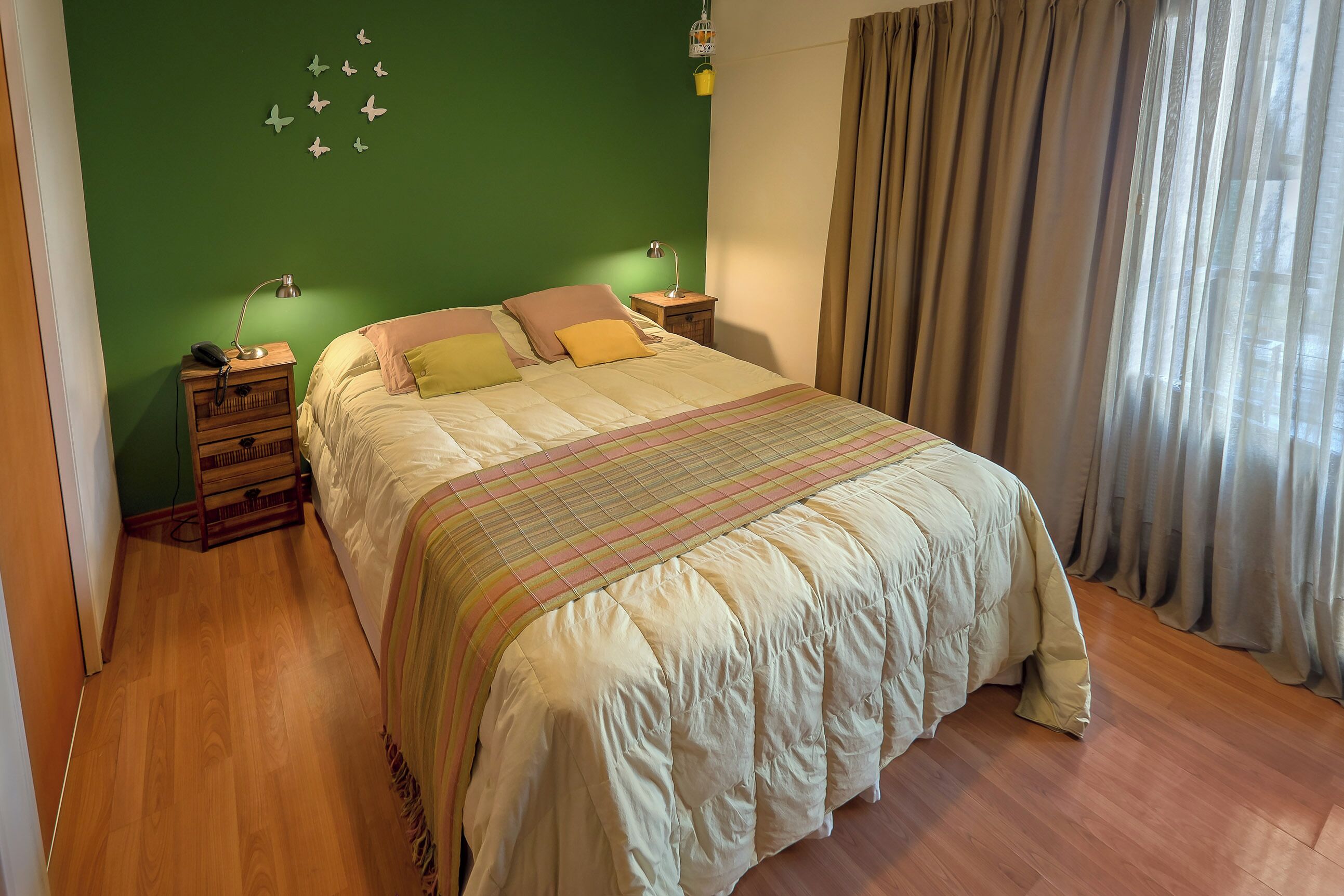 Um quarto com cama, duas mesas de cabeceira, um criado-mudo, parede verde e uma janela com cortinas.