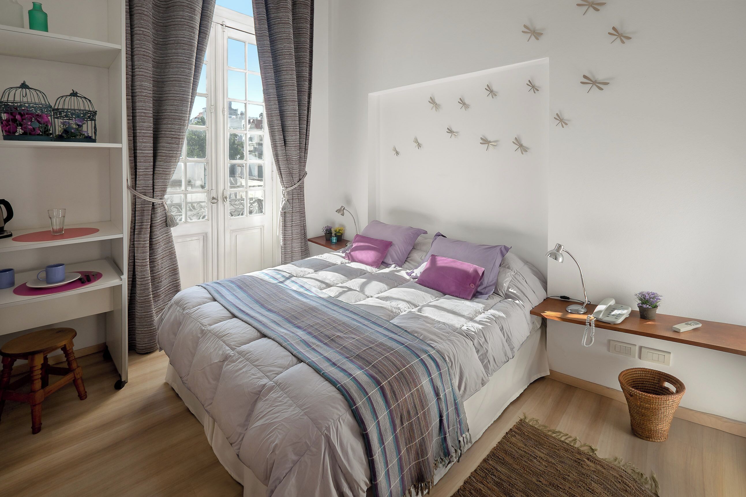 Quarto com cama, escrivaninha, cadeira, mesa de cabeceira, abajur, cesto e janela com cortinas.