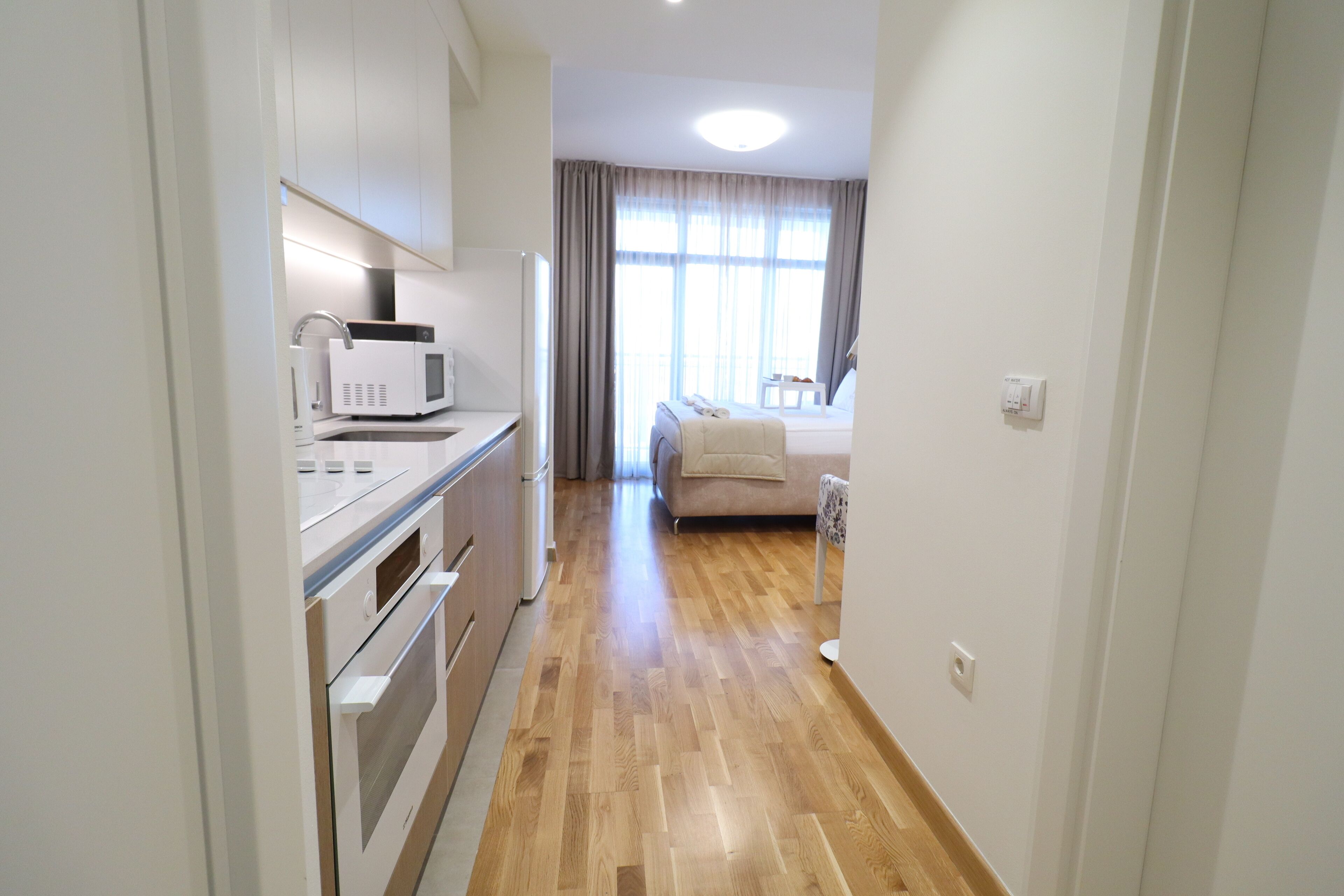 Uma cozinha moderna com eletrodomésticos brancos, piso de madeira e vista para uma sala de estar com sofá e janela.