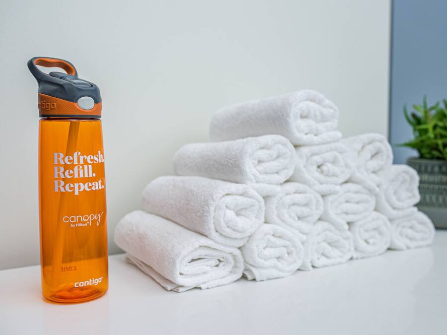 Quarto de hotel com uma pilha de toalhas brancas bem dobradas, uma garrafa de água Canopy by Hilton e uma planta em vaso.