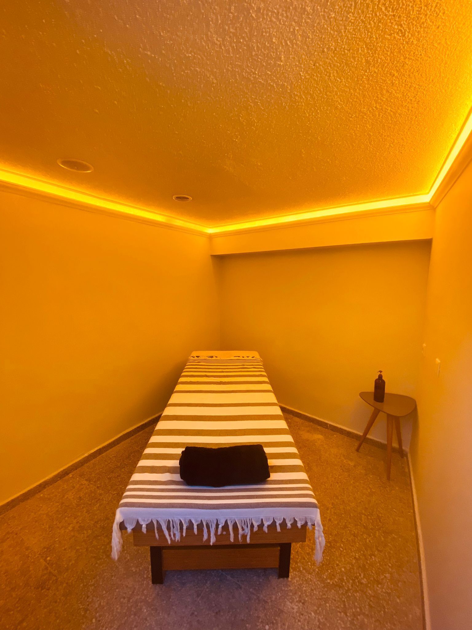 Um quarto pequeno e pouco iluminado, com uma cama de solteiro, uma mesinha e uma garrafa sobre ela.