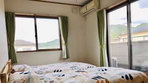Quarto com duas camas, janela com cortinas, aparelho de ar condicionado e vista para colinas.
