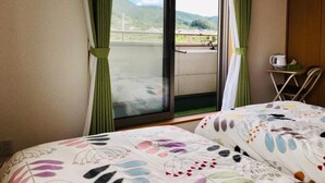 Quarto de hotel com cama, uma cadeira, uma mesinha com chaleira e vista para uma varanda com paisagem de montanha.