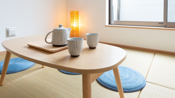 Uma mesa de madeira redonda com uma bandeja e duas xícaras de cerâmica, uma luminária alta com textura e um tapete tecido azul.