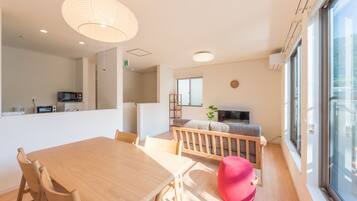Uma sala de estar moderna com uma mesa de jantar de madeira, um banquinho vermelho, um sofá cinza e uma janela grande que proporciona iluminação natural.