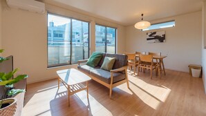 Uma sala de estar moderna com sofá, mesa de jantar e cadeiras. Janelas amplas oferecem iluminação natural e vista para prédios.