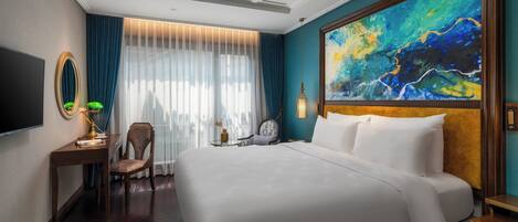 Quarto de hotel com uma cama grande, uma escrivaninha com luminária, uma cadeira, um quadro na parede e uma televisão.