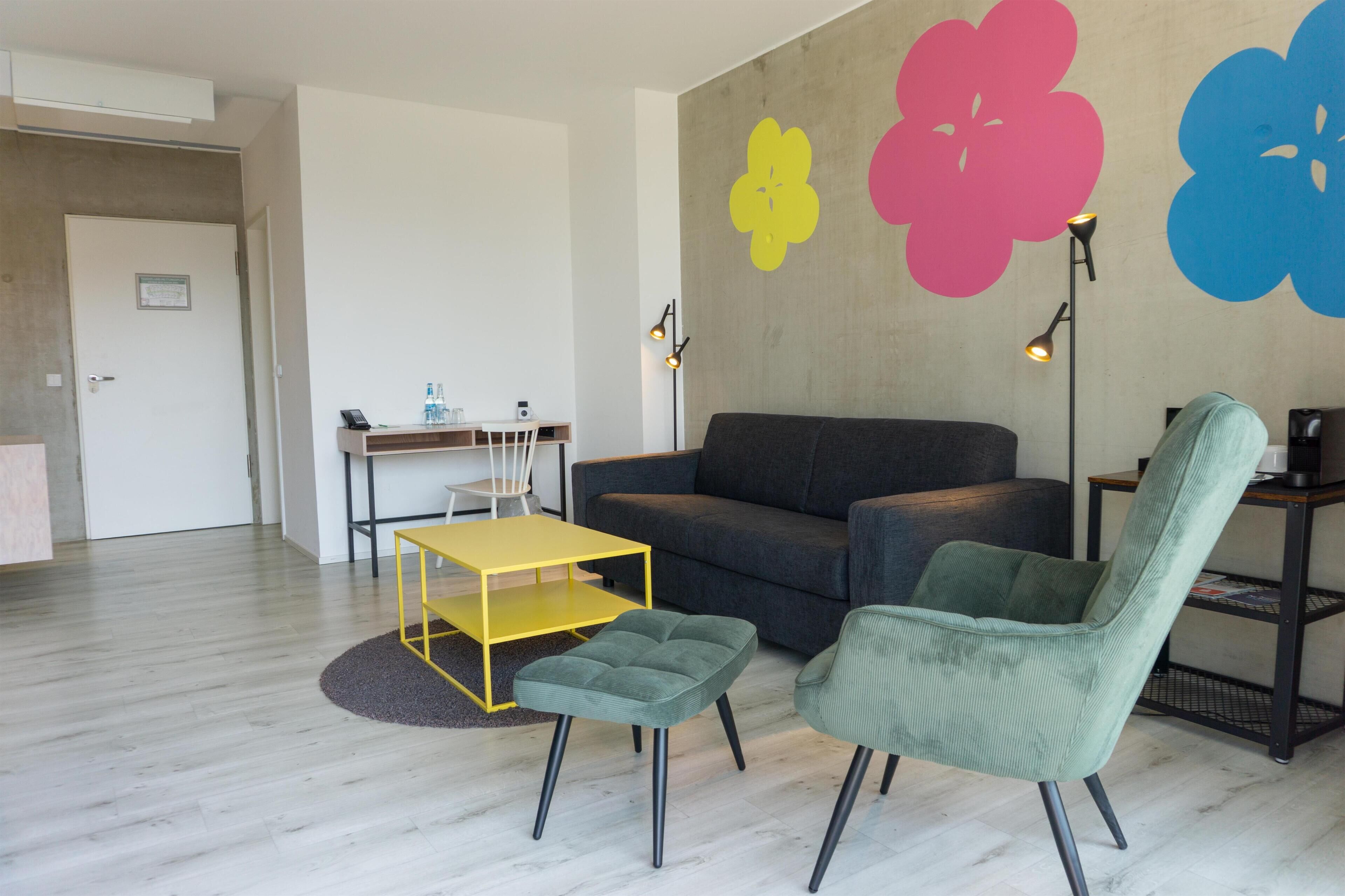 Uma sala de estar moderna com um sofá, uma poltrona verde, uma mesa de centro amarela e uma escrivaninha com cadeira.