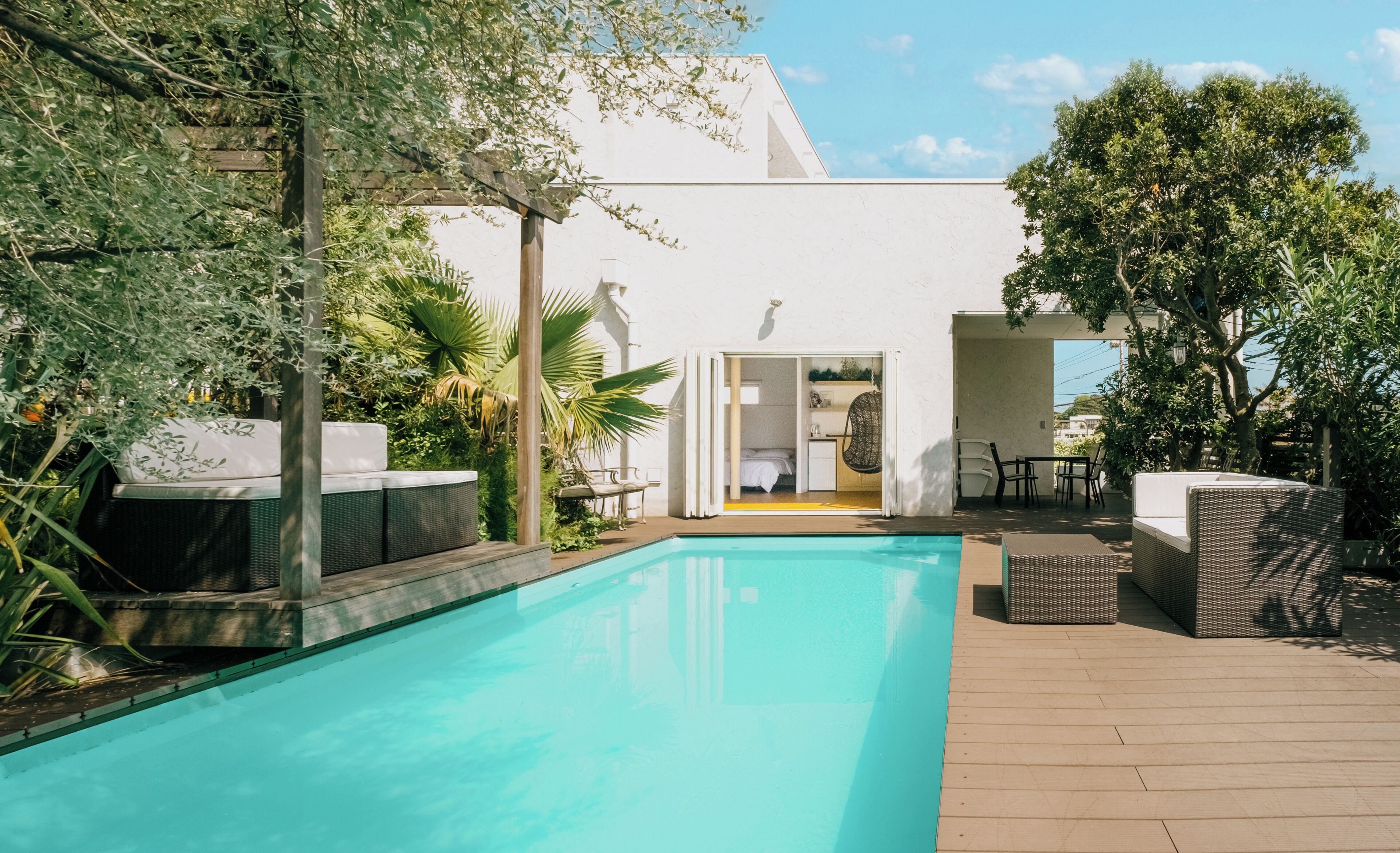 Piscina com deck de madeira, rodeada por vegetação e um edifício branco com uma porta aberta dando para um quarto.