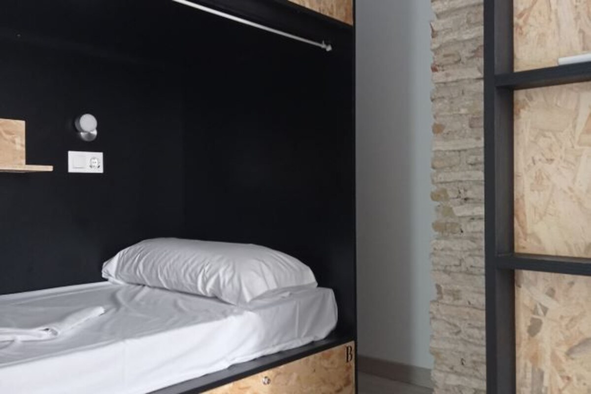 Uma cama beliche com roupa de cama branca, uma prateleira pequena com uma moldura e uma luminária fixada na parede.