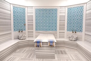 Quarto com piso de mármore, uma mesa central e duas pias fixadas na parede com padrões geométricos em azulejos azuis.