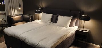Cama bem arrumada com lençóis brancos, duas luminárias laterais e cabeceira escura.