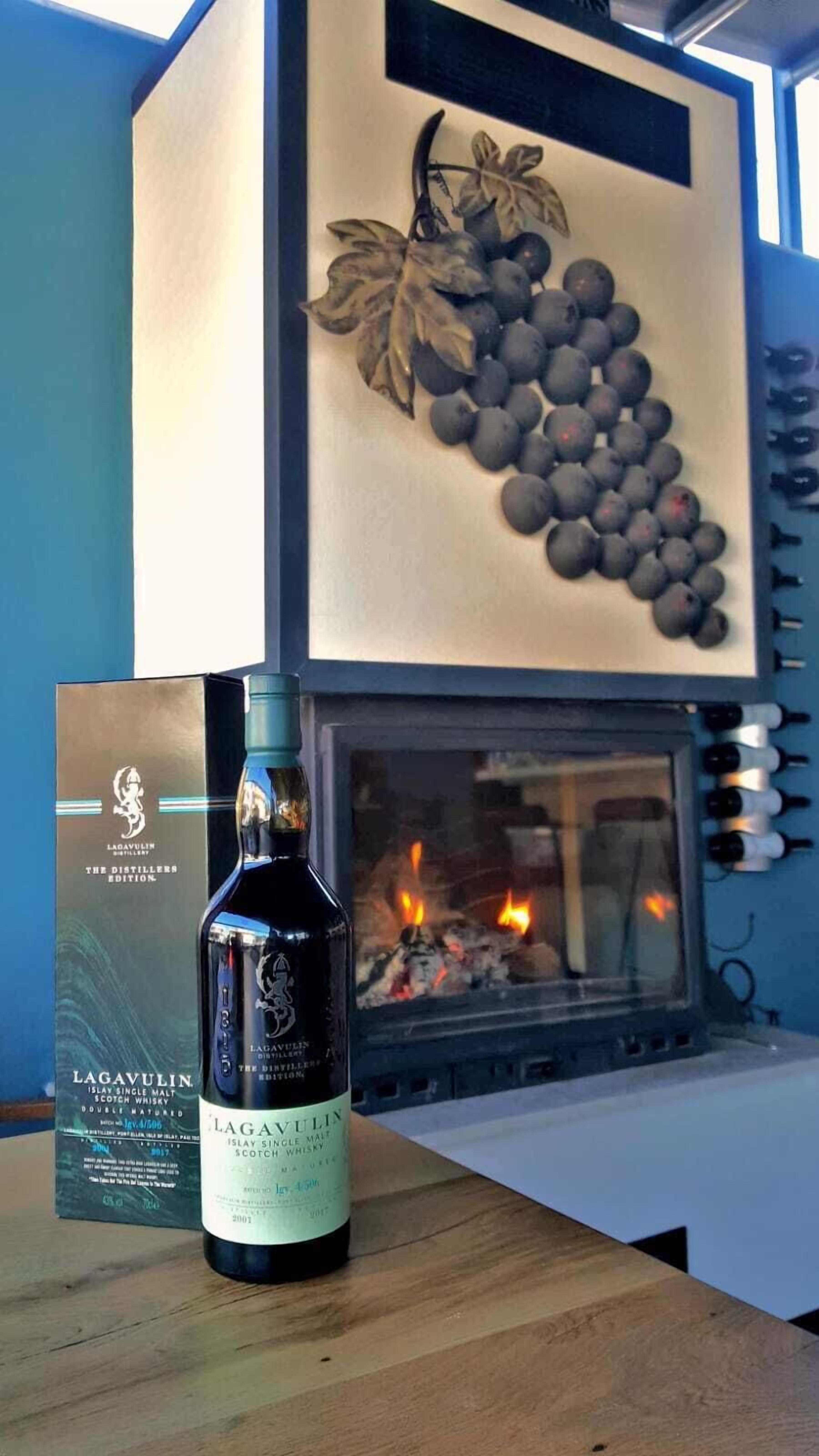 Uma garrafa de Lagavulin Islay Single Malt Scotch Whisky sobre uma mesa, em frente a uma lareira, com uma obra de arte de uvas acima.