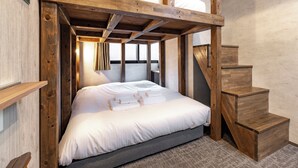 Uma cama beliche com estrutura de madeira, um colchão cinzento e uma pequena cadeira de madeira ao lado da cama.