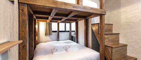 Uma cama beliche com estrutura de madeira, um colchão cinzento e uma pequena cadeira de madeira ao lado da cama.