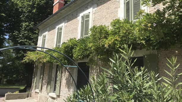 Um edifício de dois andares em tijolo com persianas verdes, uma chaminé visível e um jardim com várias plantas.